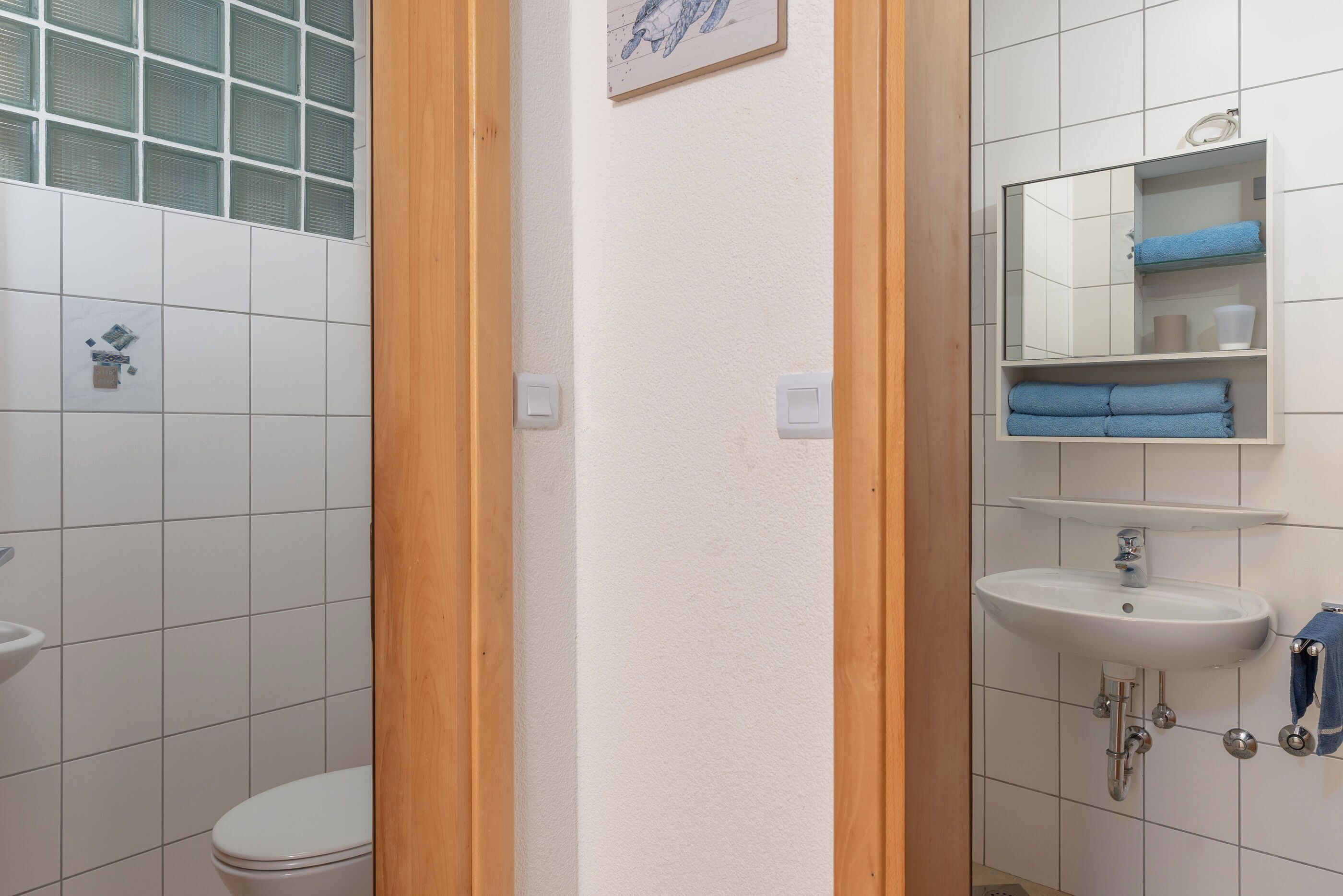 Casa de banho com azulejos brancos, sanita, lavatório e espelho com porta-toalhas.