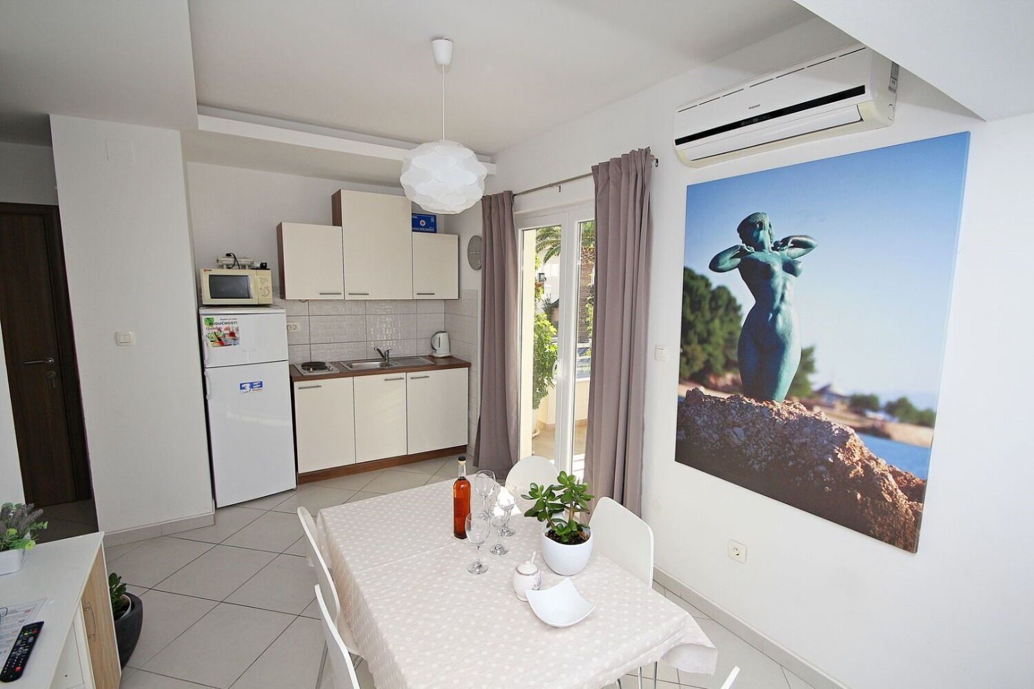 Uma cozinha compacta com eletrodomésticos modernos, um espaço de refeições com mesa posta para dois e uma grande janela com vista para uma escultura e paisagem.