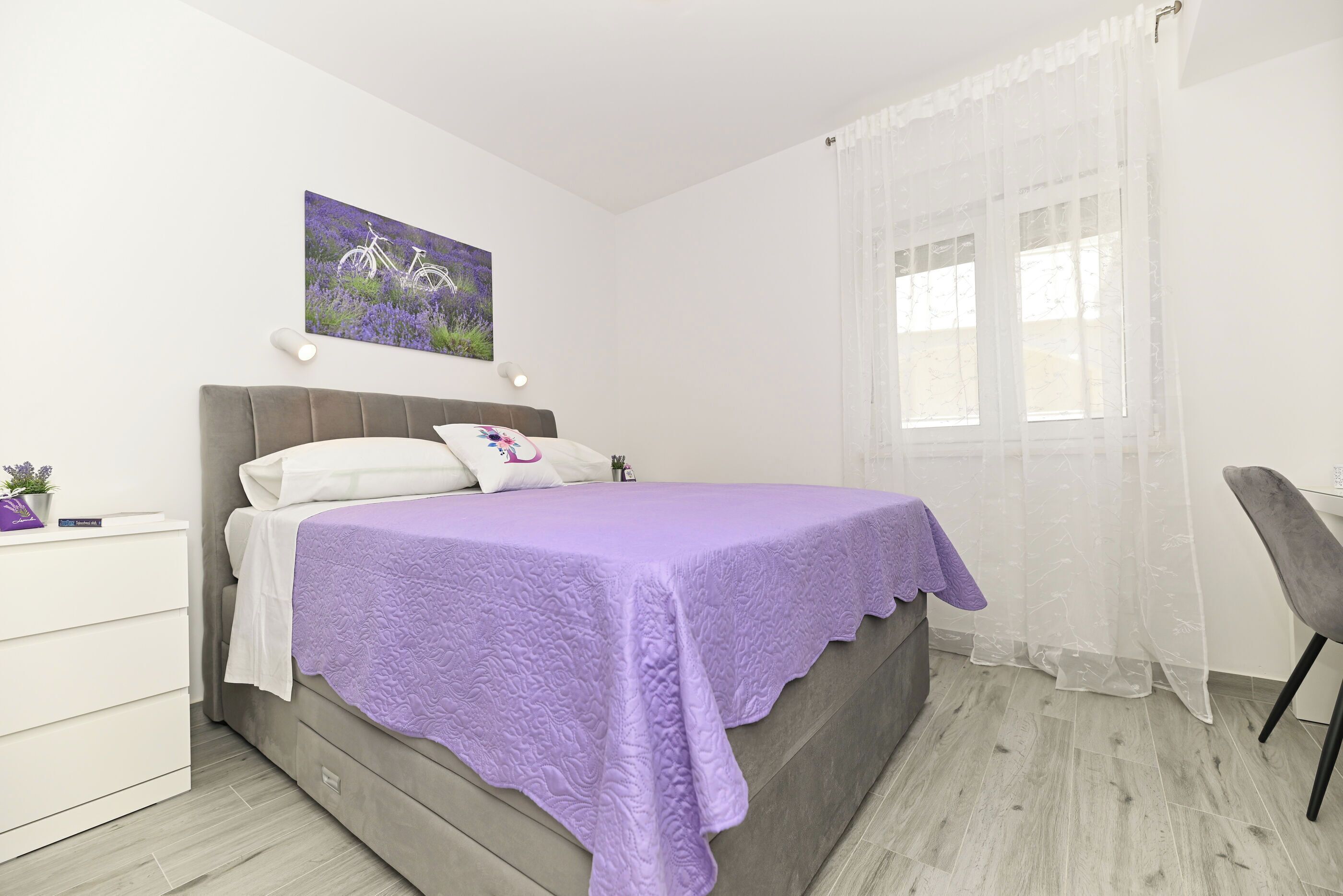 Quarto com cama, mesa de cabeceira, cadeira, janela com cortinas leves e um quadro na parede.