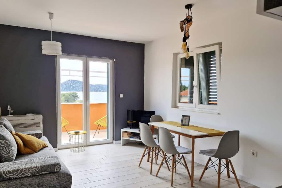 Uma sala de estar moderna com sofá, mesa de jantar e cadeiras. Há uma televisão e uma janela com persianas.