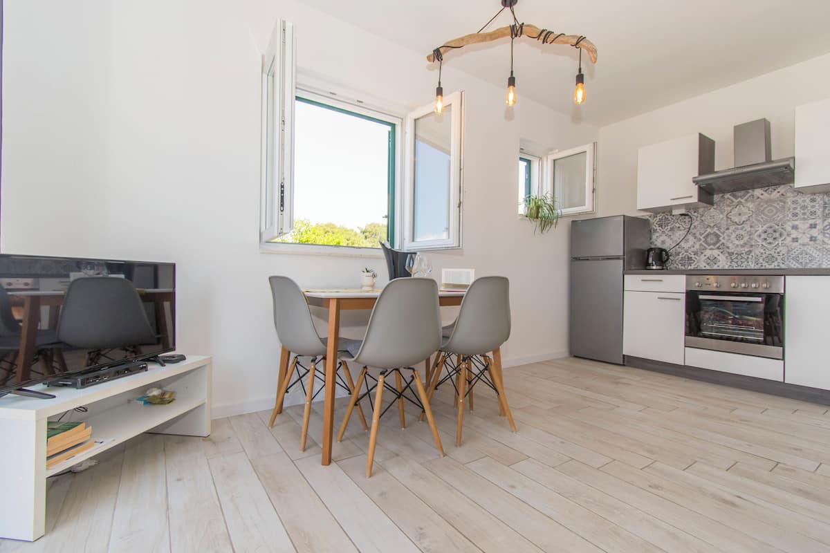 Cozinha moderna com área de jantar, apresentando uma mesa de jantar de madeira, cadeiras cinzas, forno embutido e uma janela grande com vista para o verde.