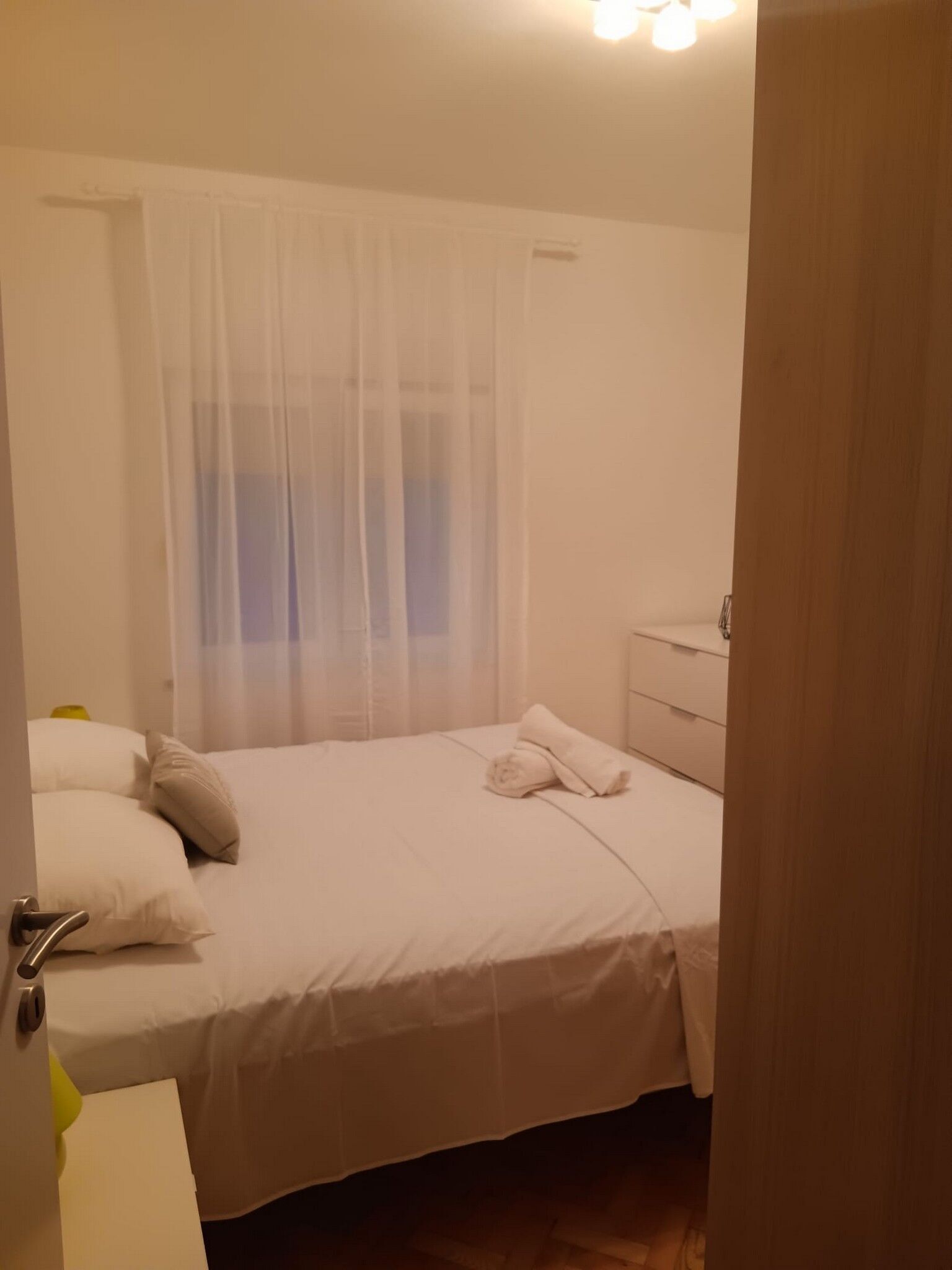 Quarto com cama, mesa de cabeceira, janela com cortinas leves e piso de madeira.