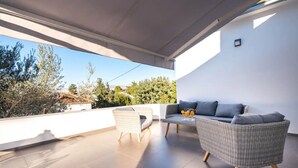 Uma varanda moderna com um sofá cinzento e cadeiras de vime, uma mesa com bebidas e vista para árvores e edifícios.