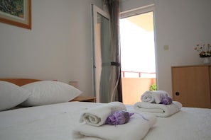 Quarto de hotel com cama, almofadas, janela com cortinas, porta e uma pequena mesa com um vaso de flores.
