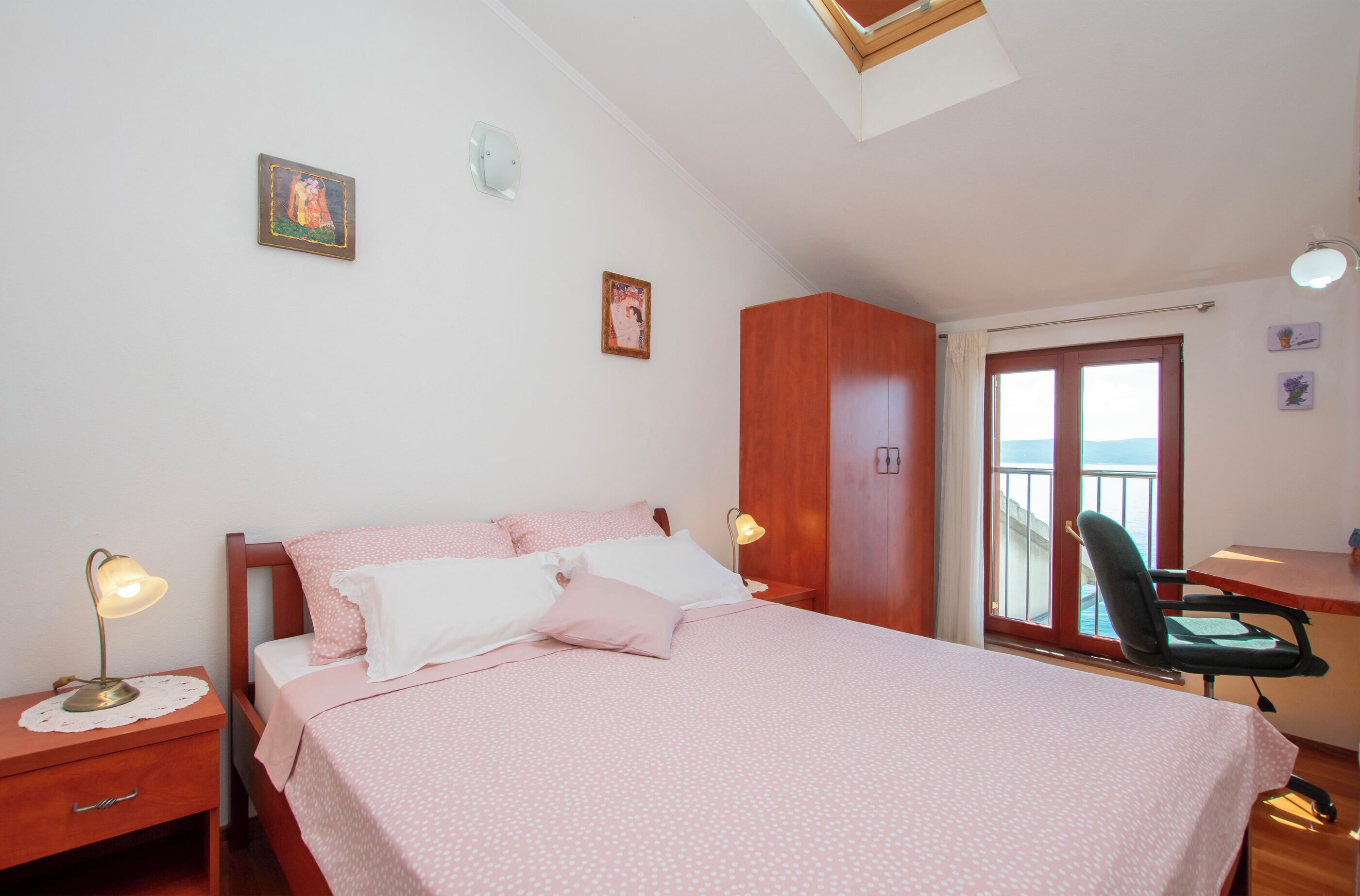 Quarto com cama, mesas de cabeceira, escrivaninha e cadeira. Há uma janela com vista para o exterior.