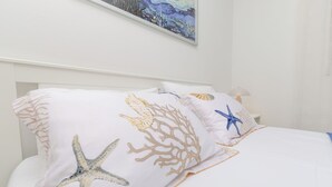 Uma cama com roupa de cama branca, estampas de estrelas-do-mar e corais, um quadro acima da cama e uma mesa de cabeceira com abajur.