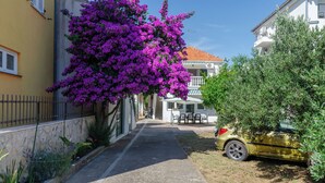 Uma rua estreita com um edifício amarelo à esquerda, um edifício branco com varanda à direita e um carro estacionado no lado direito.