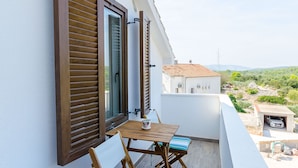 Varanda com mesa e cadeiras, janela com persianas de madeira, e vista para uma área residencial e colinas ao longe.