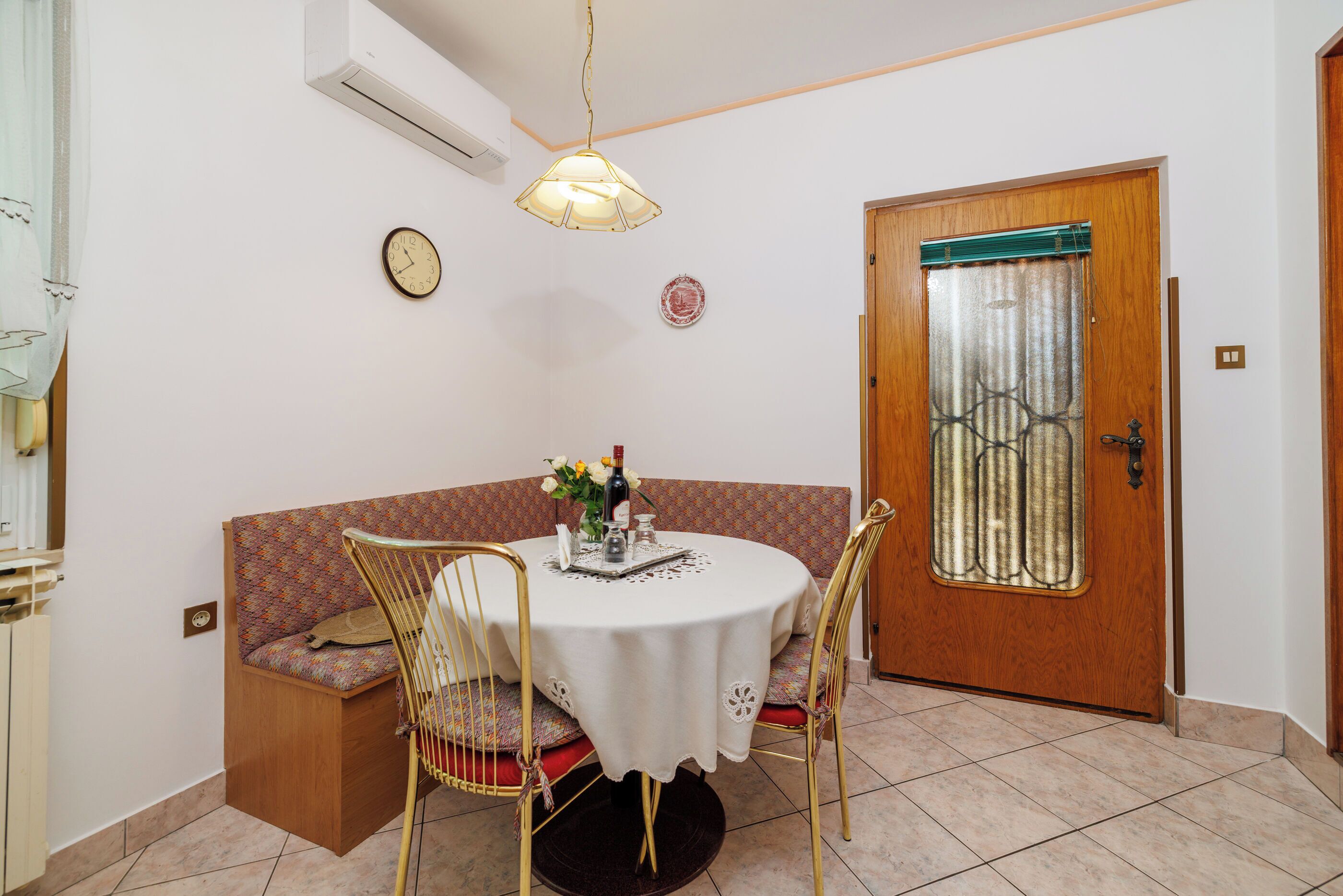 Uma sala de jantar com uma mesa redonda, cadeiras, um banco, um relógio e uma porta com cortina.