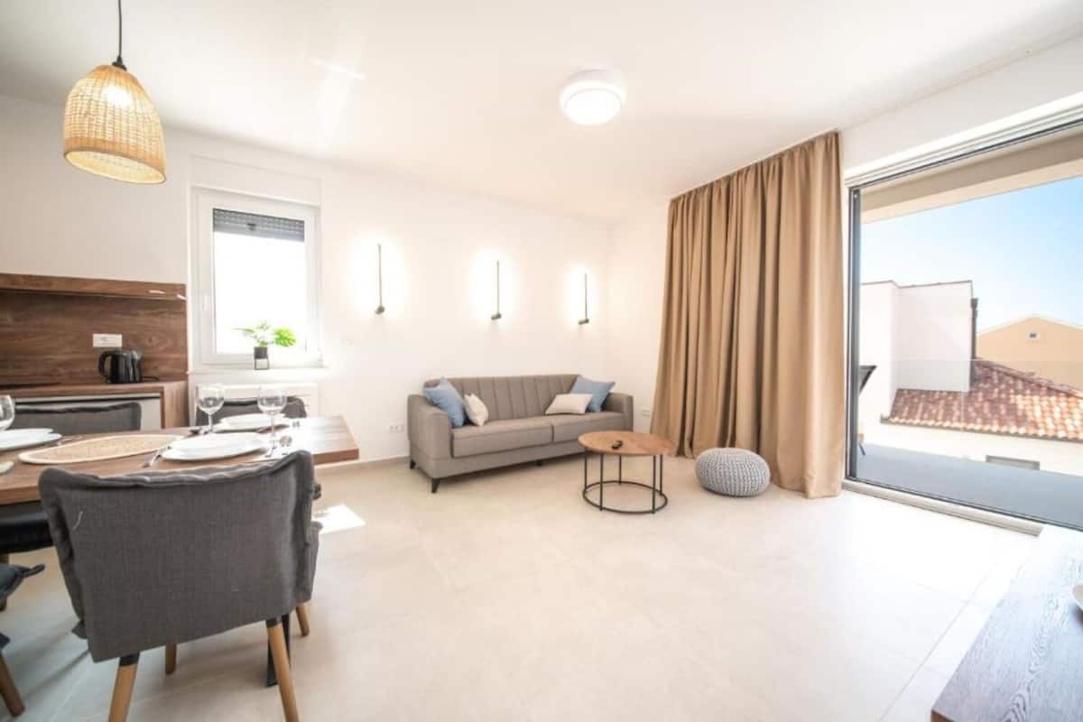 Uma sala de estar moderna com um sofá, mesa de jantar e cadeiras. Há uma janela com cortinas e vista para o exterior.