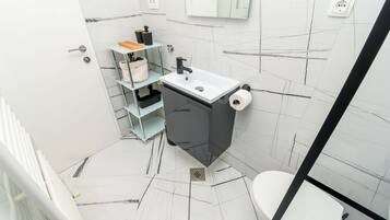 Casa de banho compacta com lavatório branco, armário preto e sanita. Existem prateleiras com produtos de higiene e um espelho sobre o lavatório.