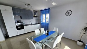 Cozinha moderna com armários brancos, forno embutido e pia. Há uma área de jantar com mesa de vidro e cadeiras brancas. Um relógio está na parede e uma planta em um canto.