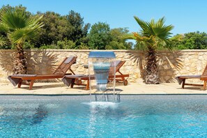 Piscina com um elemento aquático, rodeada por espreguiçadeiras e palmeiras.