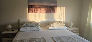 Quarto de hotel com uma cama grande, duas mesas de cabeceira, uma luminária e uma pintura de uma cena praiana na parede.