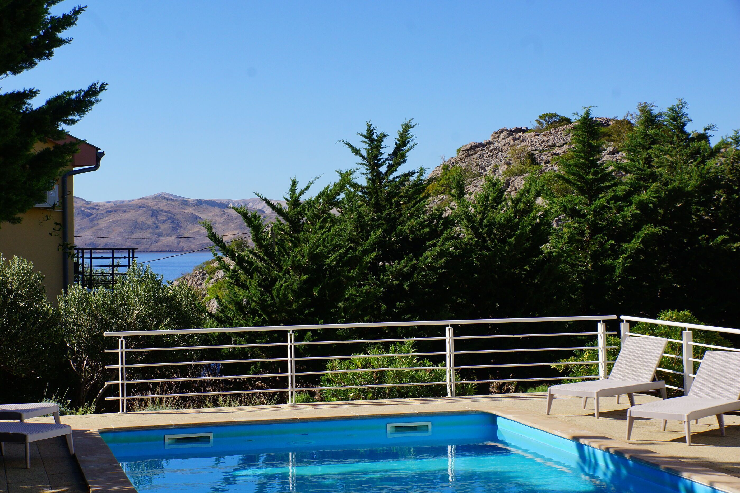 Piscina com grade de metal, duas espreguiçadeiras e vista para o mar e montanhas ao fundo.