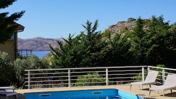 Piscina com grade de metal, duas espreguiçadeiras e vista para o mar e montanhas ao fundo.