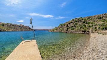 Vista nítida de uma praia de pedras que se estende até águas calmas, com uma escultura de metal à esquerda e um morro rochoso à direita.