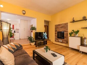 Sala de estar com sofá, mesa de centro, televisão e plantas. Cozinha visível ao fundo.