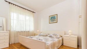 Quarto com cama, mesas de cabeceira, cômoda, janela com cortinas leves e um quadro na parede.