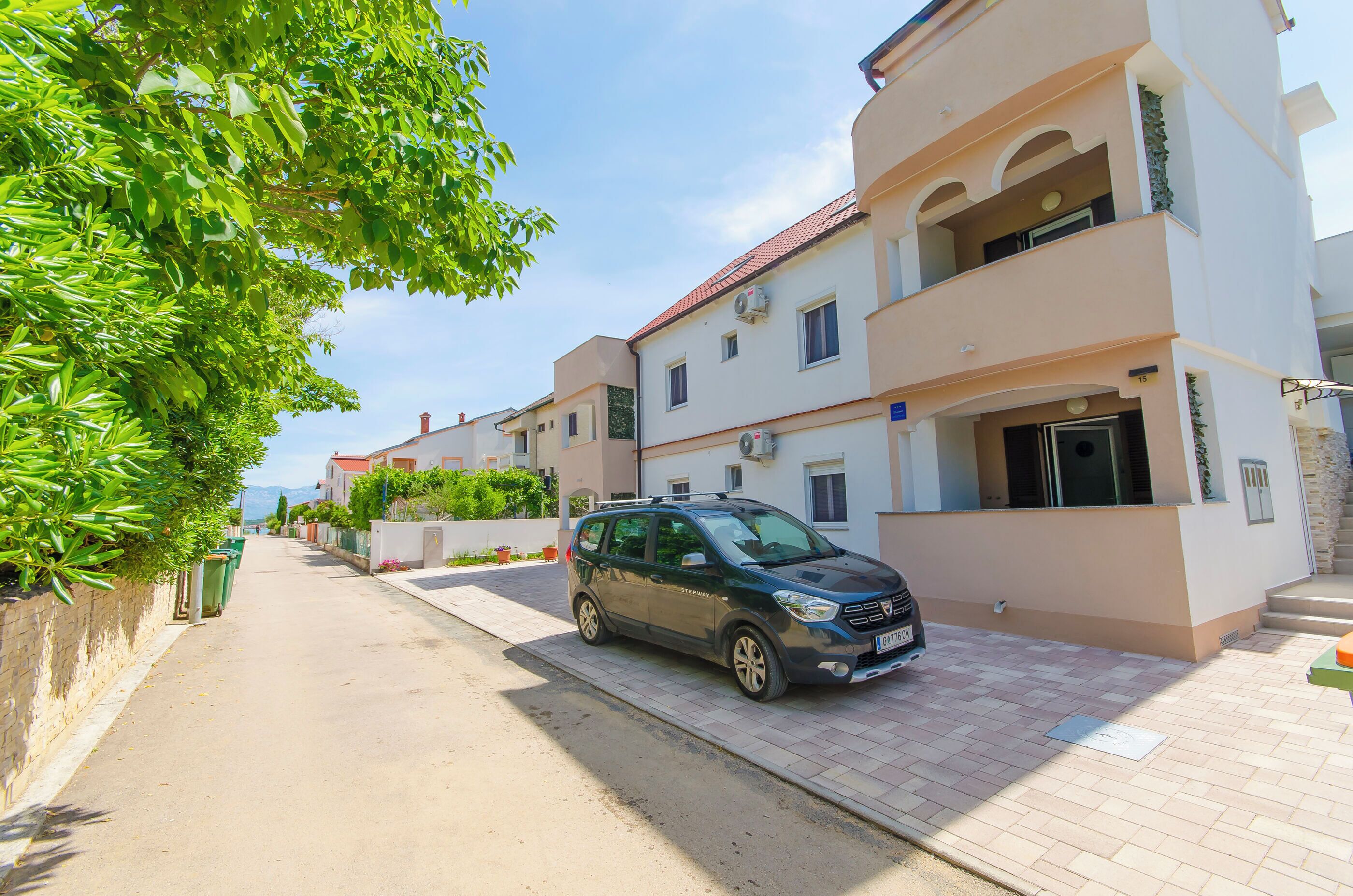 Uma rua residencial com um carro estacionado, casas com sacadas e um céu claro.