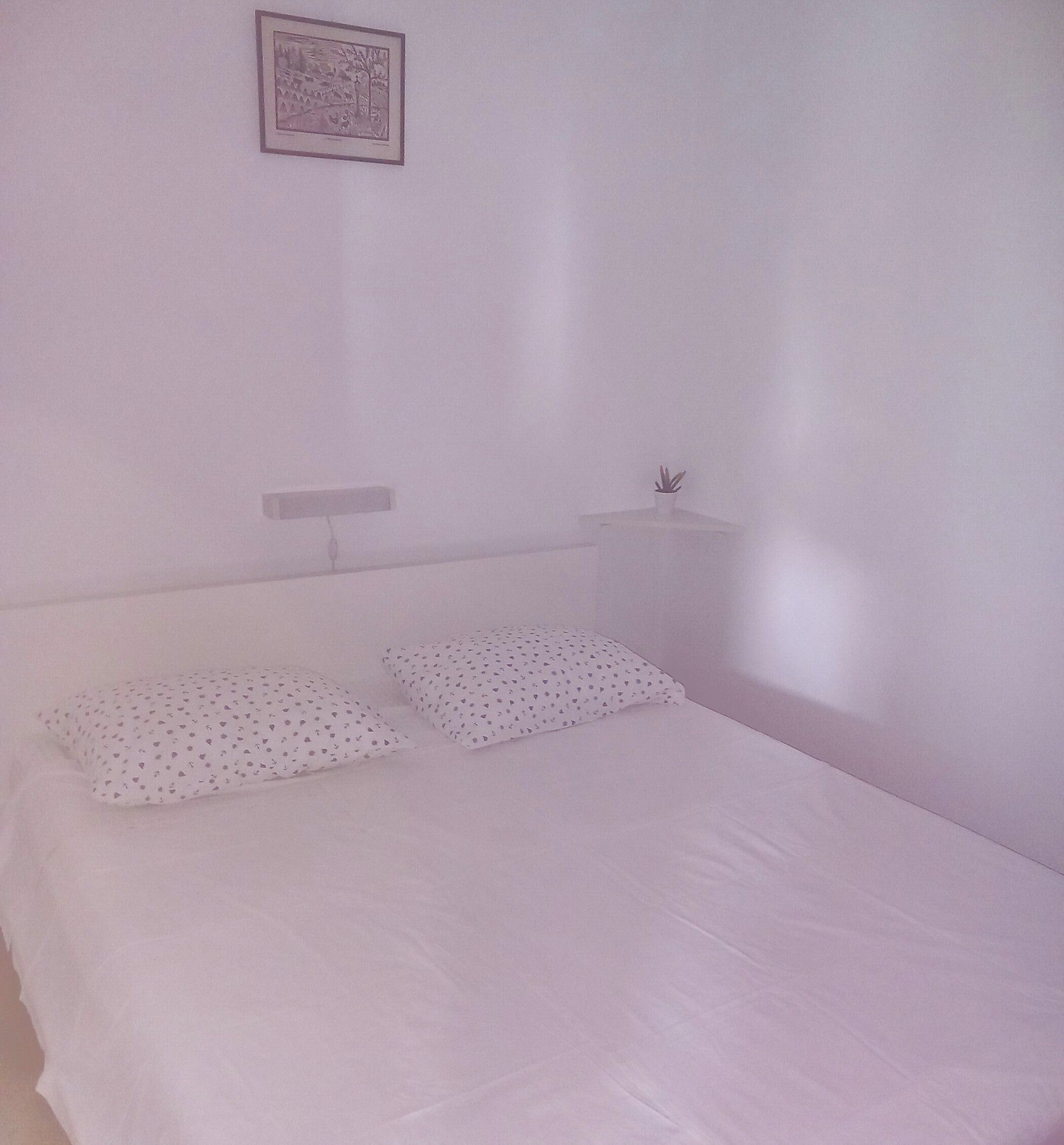 Quarto simples e claro com uma cama, duas almofadas, uma pequena mesa de cabeceira com uma planta e um quadro na parede.