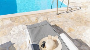 Uma cadeira de lounge com almofada, chapéu de palha, óculos de sol e toalha de praia junto a uma piscina com piso de pedra.