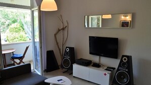 Uma sala de estar moderna com televisão de tela plana, dois alto-falantes grandes, um sofá, uma mesa pequena e uma janela com vista para o verde.