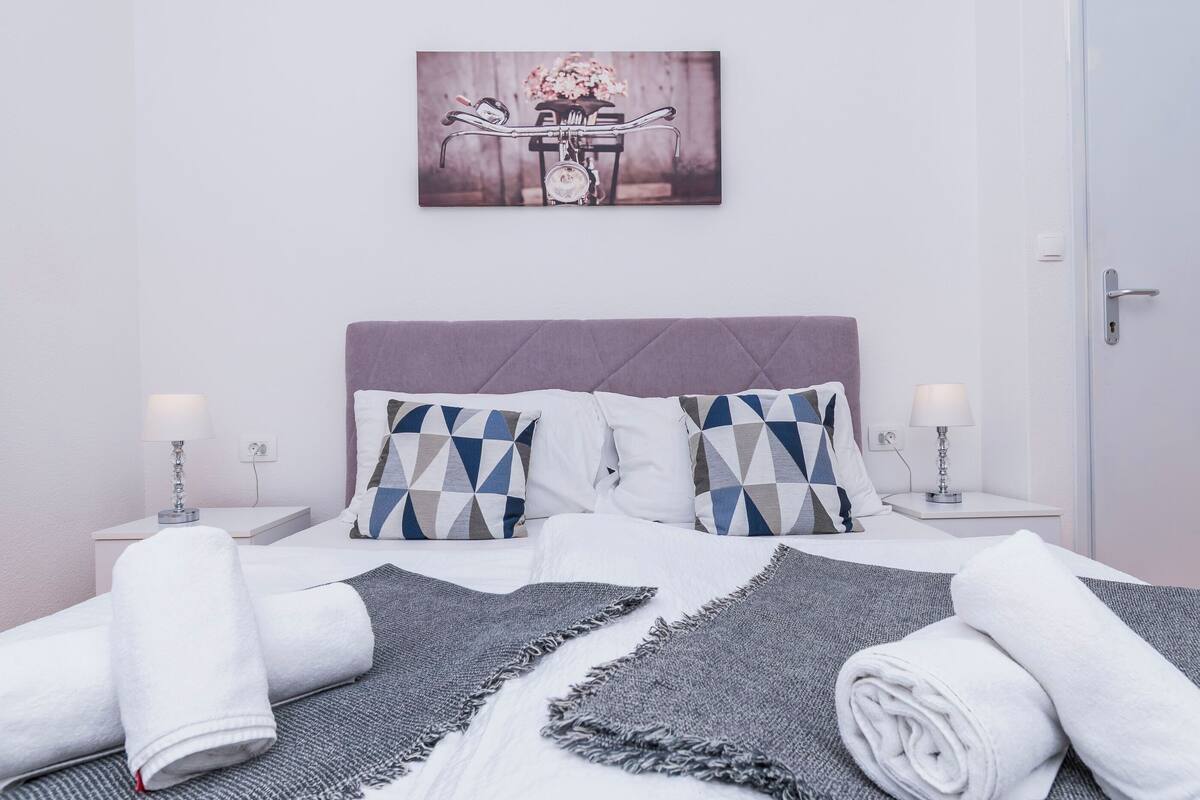 Cama bem arrumada com manta cinza e toalhas brancas dobradas, dois travesseiros estampados e um quadro pendurado na parede acima da cama.