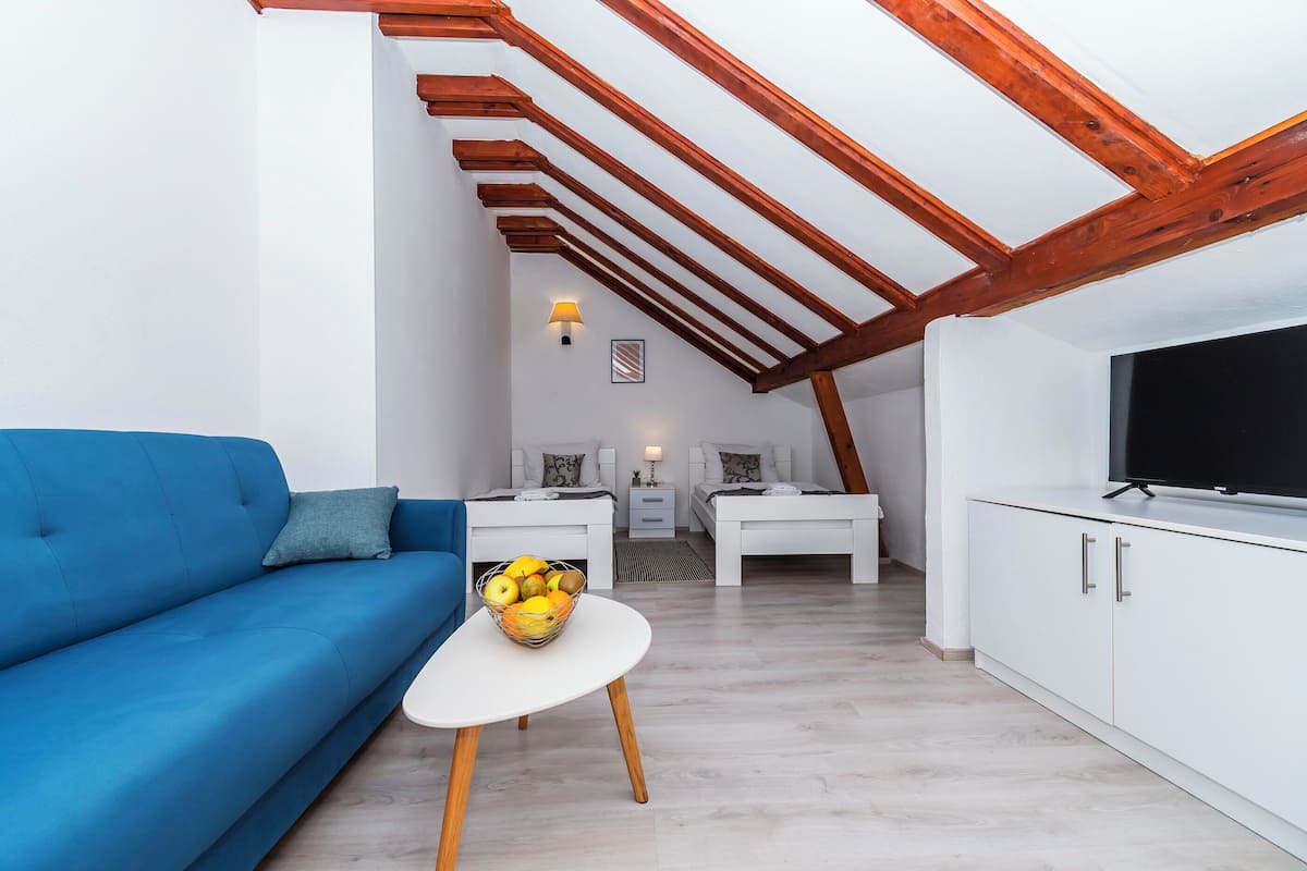 Um quarto moderno no estilo loft, com teto inclinado, um sofá azul, uma mesa branca com uma cesta de frutas, uma TV em um móvel e uma pequena mesa de cabeceira com uma luminária.