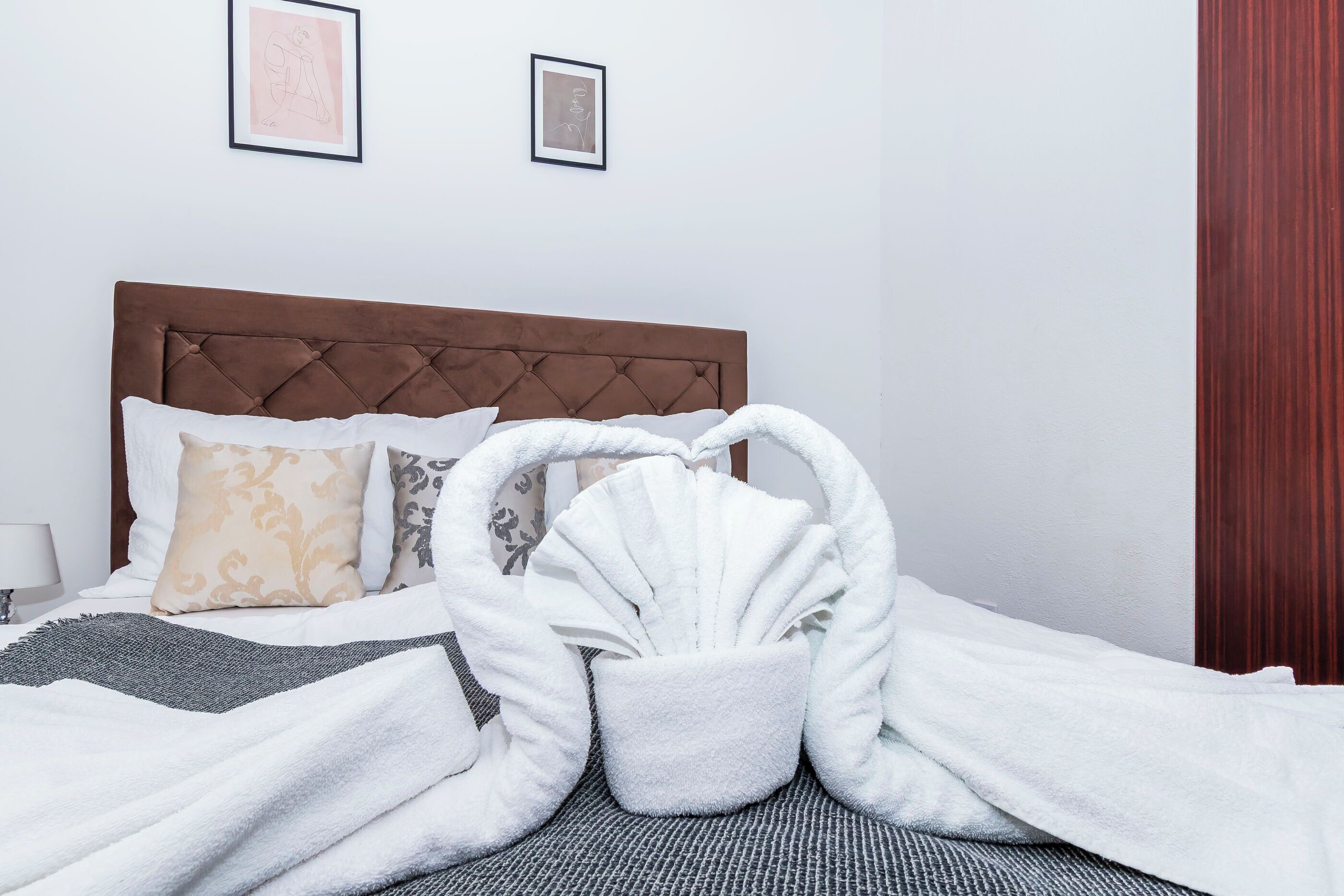 Cama bem arrumada com cabeceira marrom, almofadas brancas e estampadas, e toalha dobrada em forma de coração.