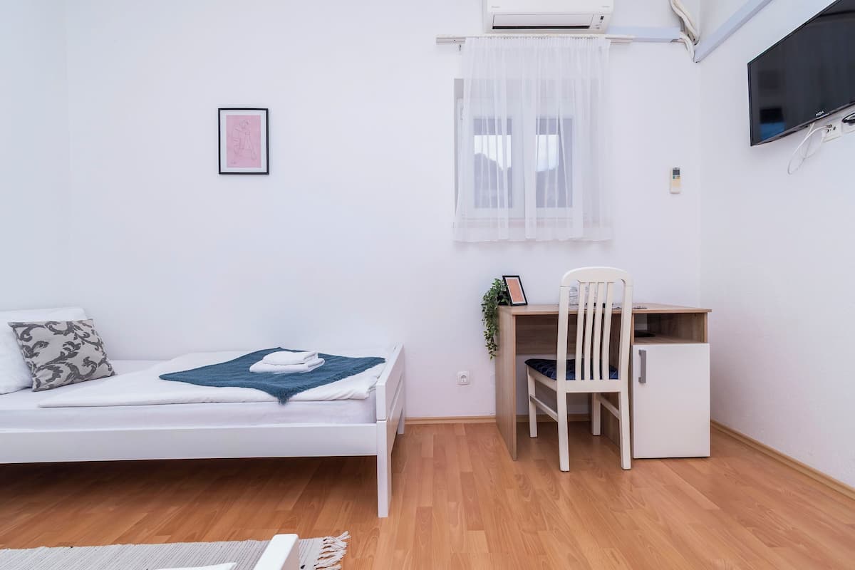 Quarto minimalista com cama branca, escrivaninha com cadeira, TV na parede e janela com cortinas leves.