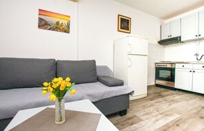 Uma sala de estar com um sofá cinza, uma mesa pequena com um vaso de flores amarelas, uma geladeira branca e uma cozinha com armários brancos e um fogão.