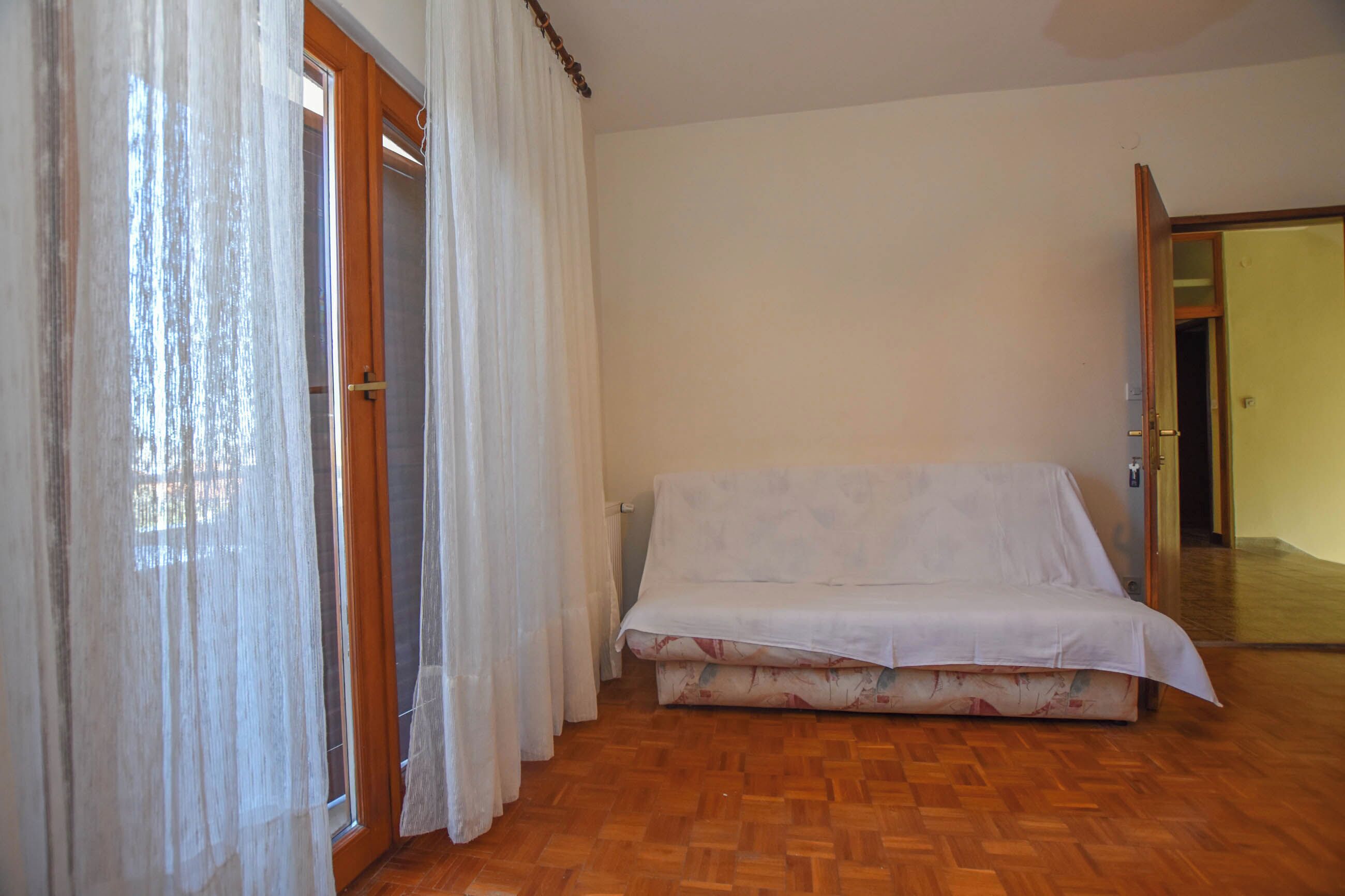Quarto com chão de madeira, cama com cobertura floral e cortinas brancas.
