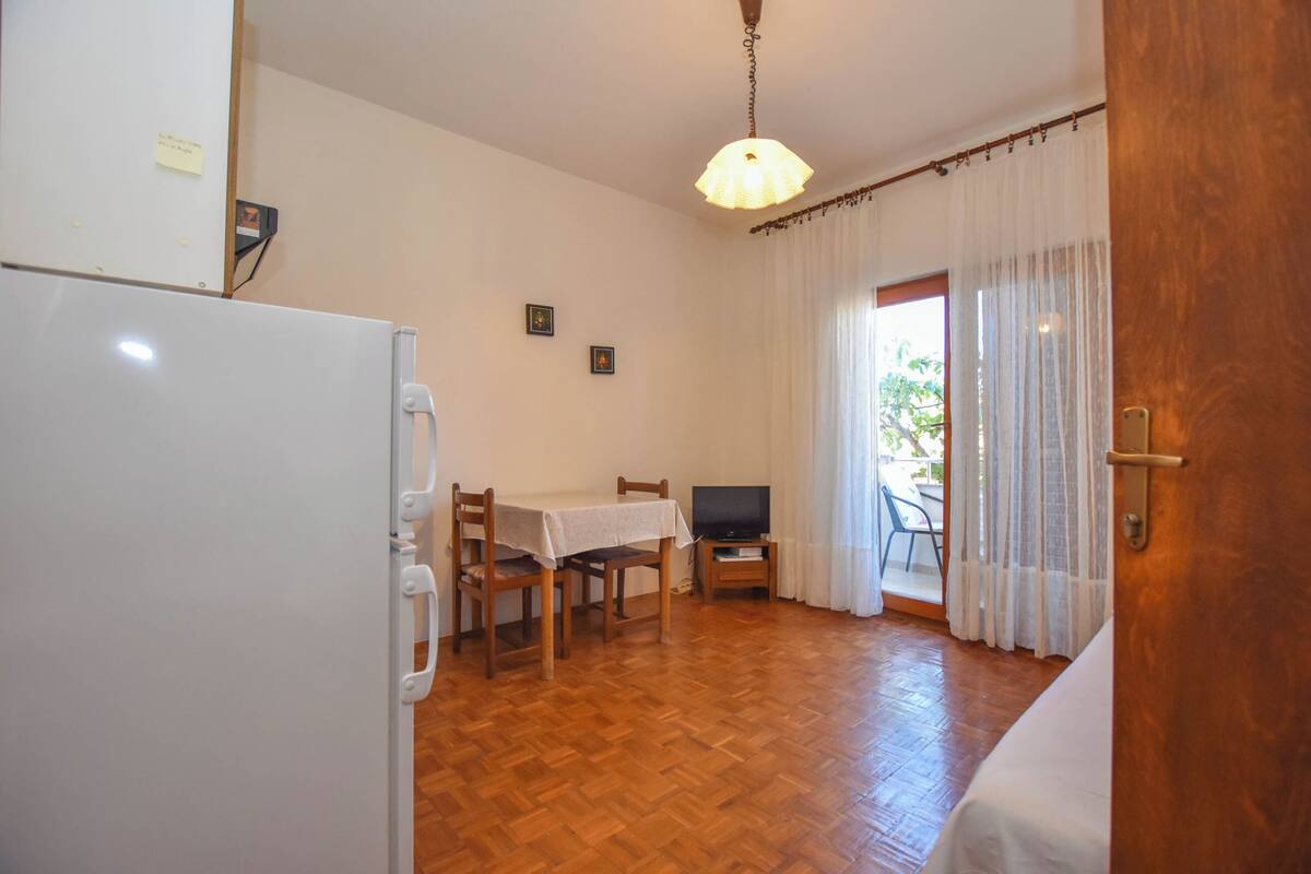 Quarto compacto com piso de madeira, mesa de jantar com cadeiras, televisão e geladeira.