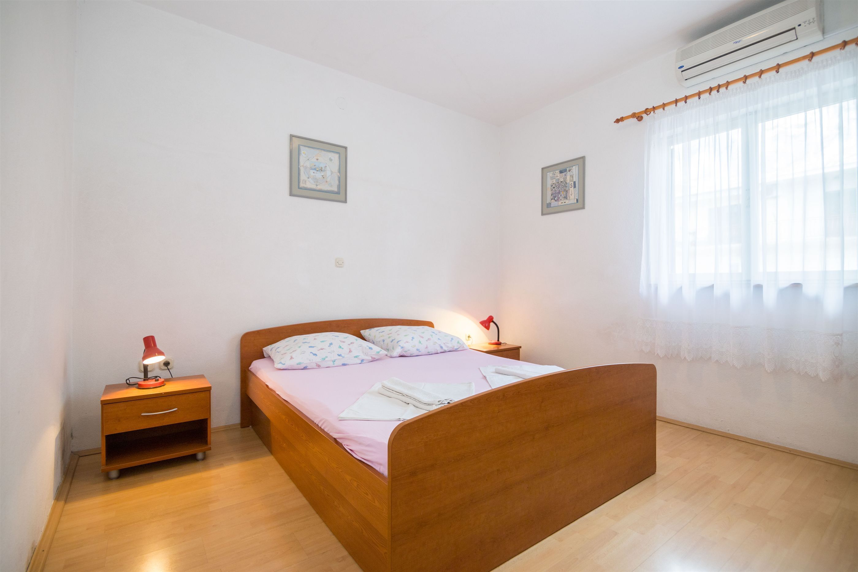 Quarto com cama de madeira, mesa de cabeceira com abajur e janela com cortinas leves.