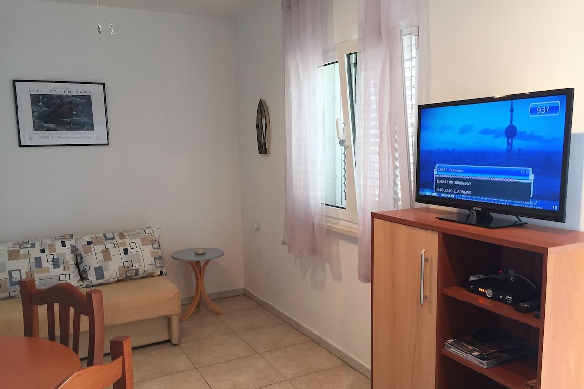 Uma sala de estar com televisão exibindo um noticiário, um sofá, uma mesinha e um armário.