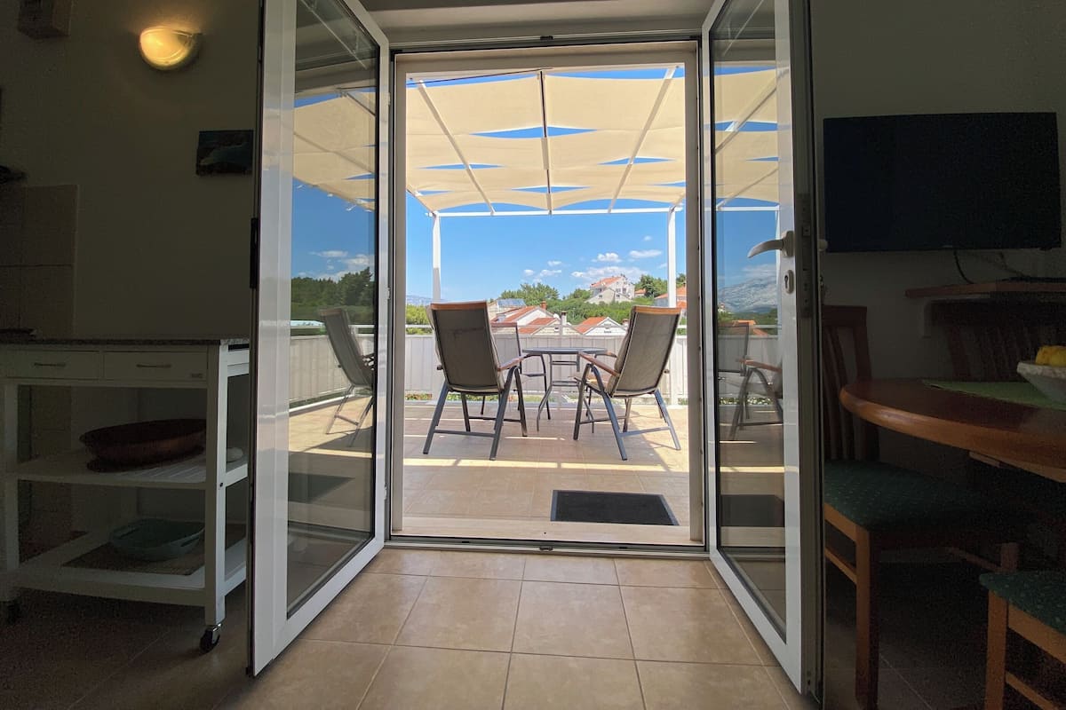 Quarto com porta de correr de vidro que dá para um espaço de pátio ao ar livre, com mesa e cadeiras, TV na parede e cozinha com armários e bancada.