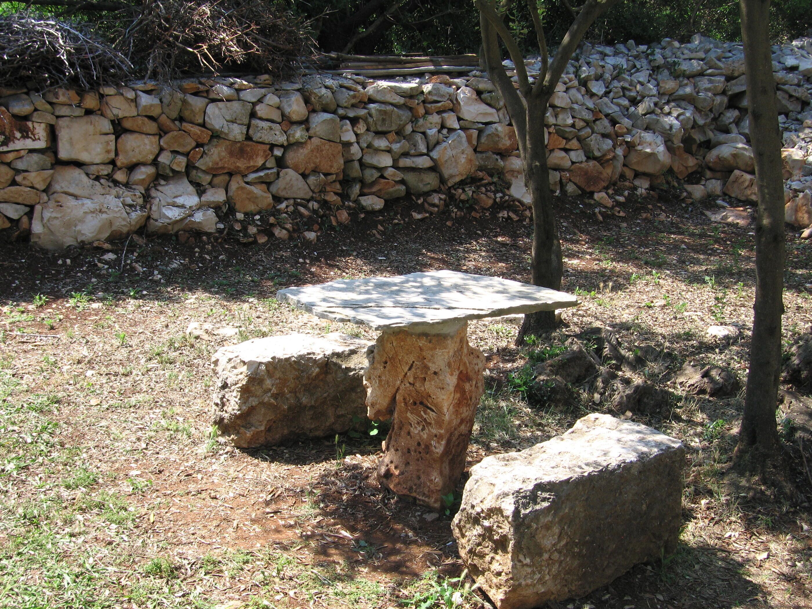 Um muro de pedra, uma mesa de pedra e duas pedras grandes em um cenário natural.