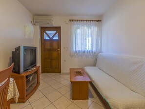 Uma sala de estar pequena com televisão sobre um móvel de madeira, um sofá, uma mesinha e uma janela com cortinas leves.