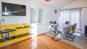 Uma sala de estar moderna com uma TV de tela plana na parede, piso de madeira, uma mesa de jantar com cadeiras e uma janela com cortinas.