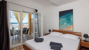 Quarto de hotel com cama, mesa de cabeceira, pintura de uma praia e vista para o mar através de uma porta de vidro.