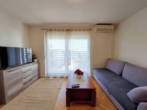 Uma sala de estar com sofá, uma televisão sobre um móvel de madeira, mesa de centro e uma janela grande com cortinas.
