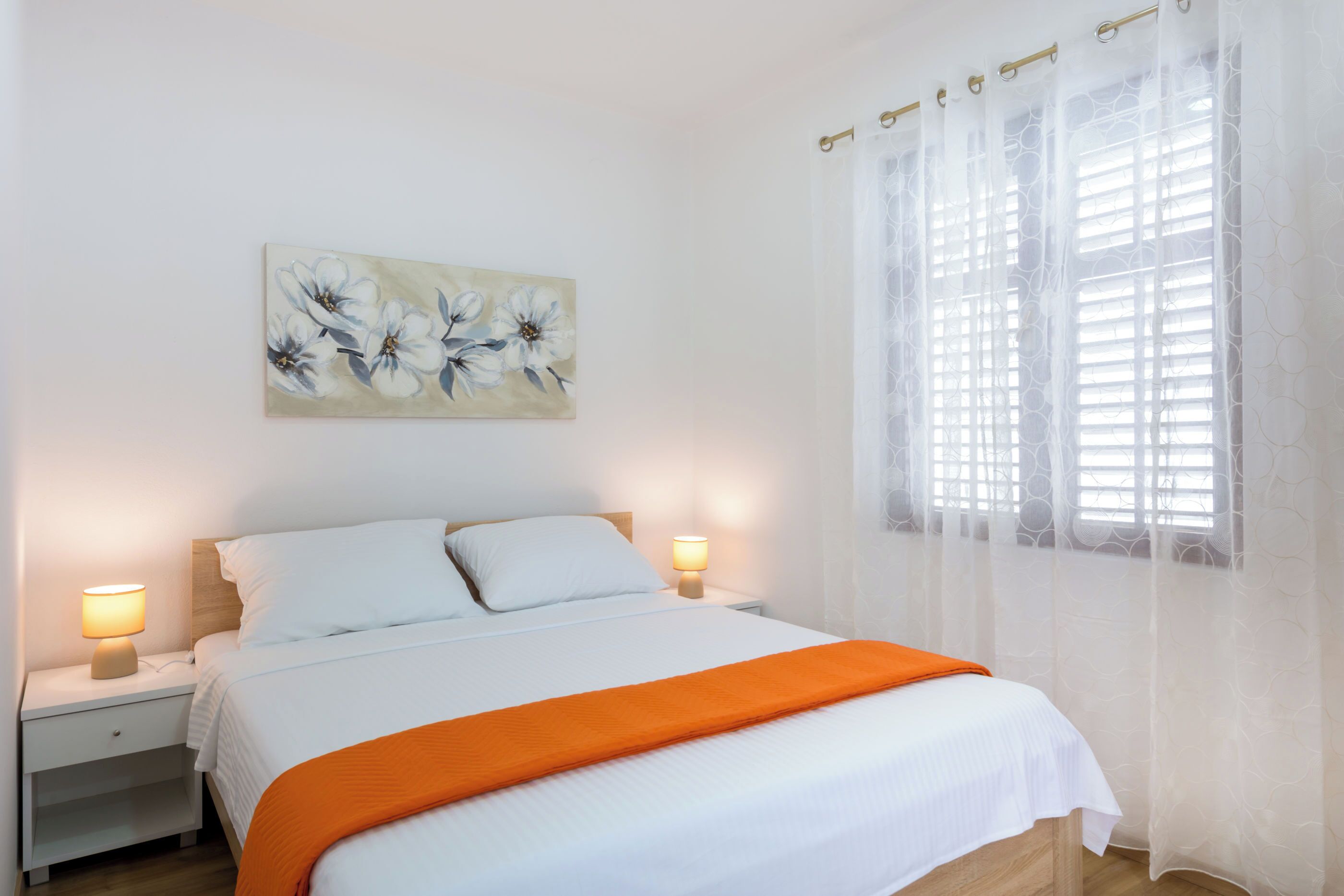Uma cama bem arrumada com roupa de cama branca e um cobertor laranja, uma luminária de cabeceira e um quadro de flores pendurado na parede.