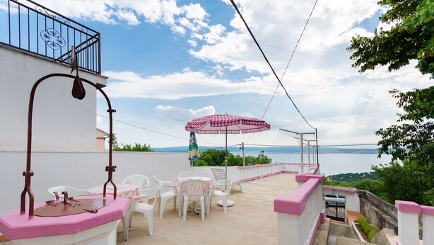 Terraço com mesa e cadeiras, um guarda-sol rosa e vista para o mar.