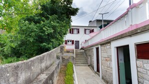 Uma passagem estreita com um muro de concreto de um lado e uma fileira de edifícios com paredes de pedra e persianas vermelhas.