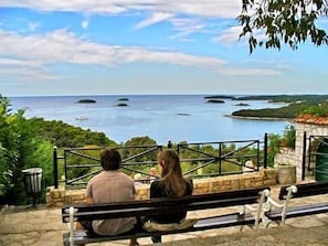 Duas pessoas sentadas num banco apreciando a vista costeira, com um muro de pedra e vegetação.
