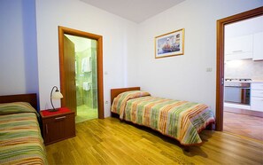 Quarto pequeno e bem iluminado, com duas camas de solteiro, uma mesa de cabeceira de madeira, uma escrivaninha com luminária, um quadro pendurado na parede e um box de chuveiro com divisória de vidro.