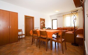 Uma sala de jantar com uma mesa posta para quatro pessoas, cercada por cadeiras de madeira. Há um guarda-roupa de madeira à esquerda e um sofá à direita.