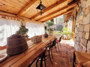 Uma mesa de madeira com cadeiras, um barril de pedra e um vaso de plantas em área externa coberta, com paredes de pedra e vista para um edifício ao fundo.