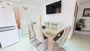 Uma sala de jantar moderna com mesa de vidro, cadeiras acolchoadas e televisão de tela plana na parede.